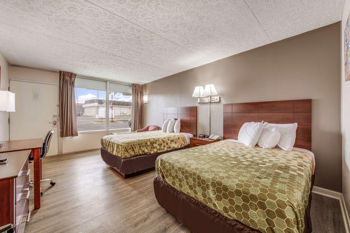 Quarto de hotel com duas camas, uma escrivaninha, uma cadeira, uma janela com cortinas e uma luminária na parede.