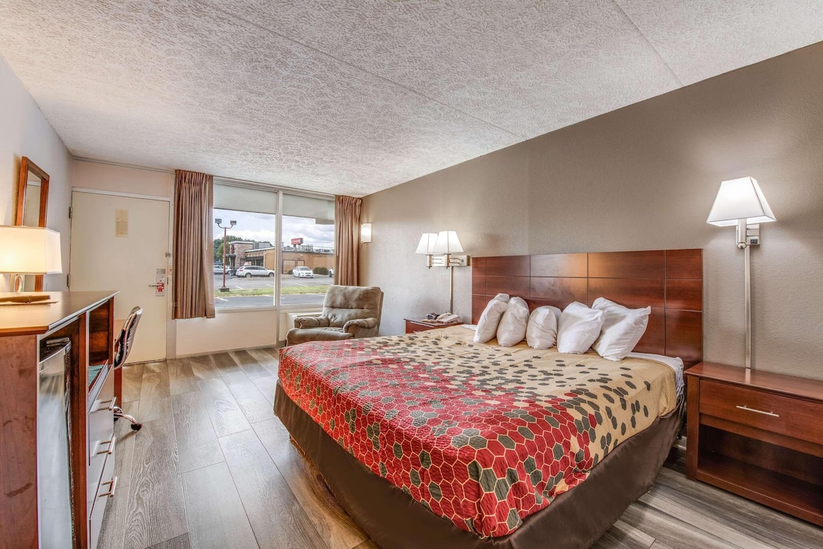 Quarto de hotel com uma cama, duas luminárias laterais, uma escrivaninha com luminária, uma cadeira e vista para o exterior.
