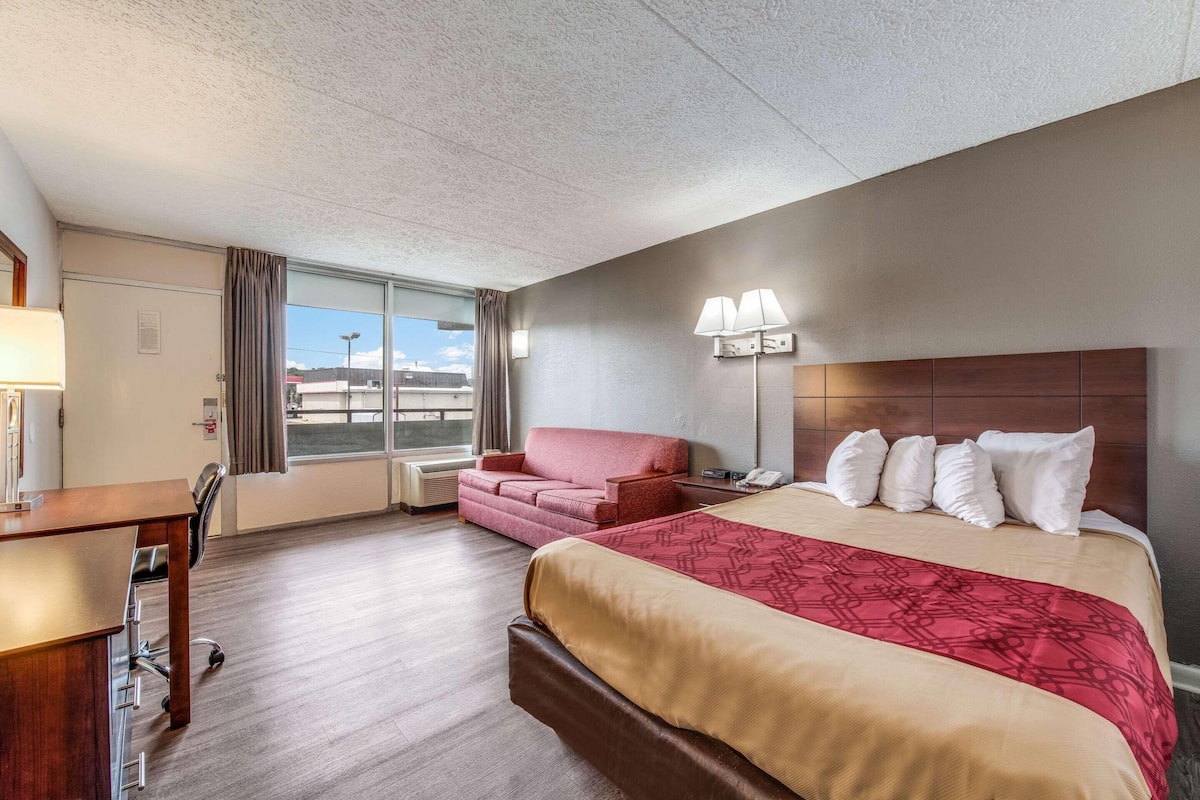 Quarto de hotel com cama, escrivaninha, cadeira, sofá, luminária e janela.