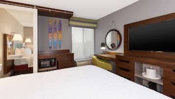 Quarto de hotel com uma cama grande, uma escrivaninha com cadeira, televisão, micro-ondas e um espaço de banheiro com pia e espelho.