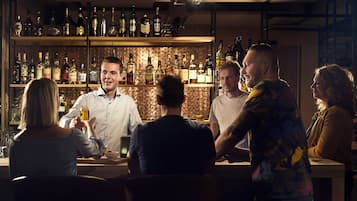 Uma área de bar com um bartender e clientes. Há prateleiras com diversos tipos de garrafas e copos.