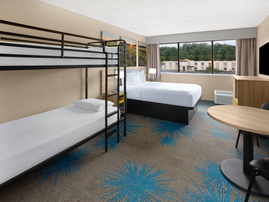 Quarto de hotel com beliches, uma cama de solteiro, escrivaninha, cadeira e uma janela grande com vista para prédios e árvores.