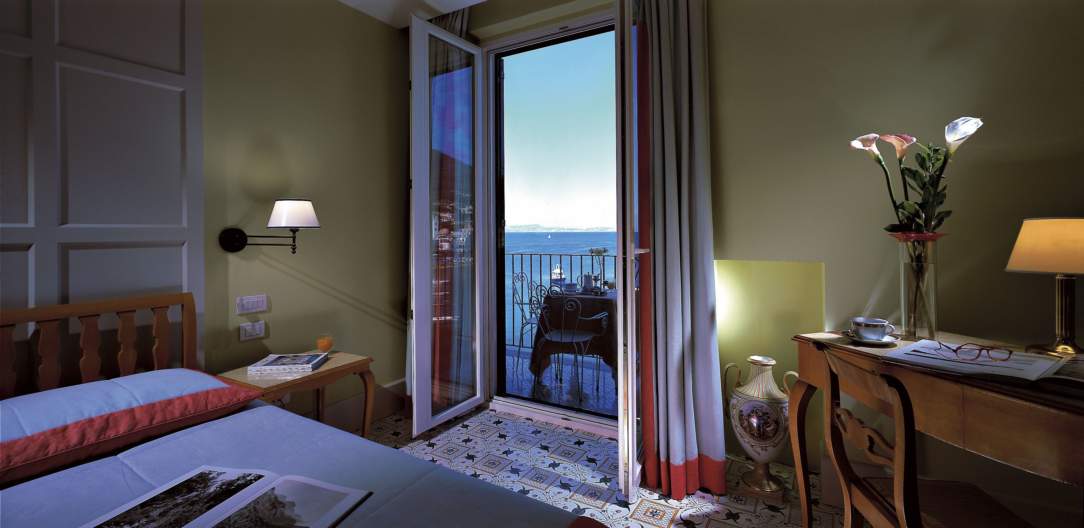 Quarto de hotel com cama, escrivaninha, cadeira, vaso com flores e vista para o mar através de uma porta de vidro.