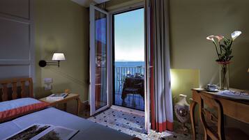 Quarto de hotel com cama, escrivaninha, cadeira, vaso com flores e vista para o mar através de uma porta de vidro.