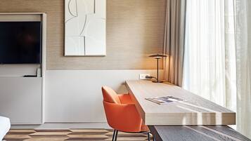 Quarto de hotel moderno com televisão de tela plana, mesa de jantar de madeira, uma cadeira laranja e uma grande janela com cortinas translúcidas.