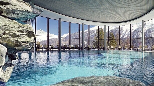 Uma piscina com água azul cristalina, rodeada por grandes janelas com vista para as montanhas.
