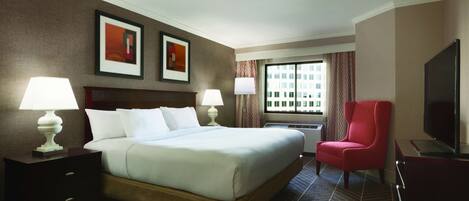 Quarto de hotel com uma cama grande, duas mesas de cabeceira com luminárias, uma poltrona vermelha, uma televisão e uma janela com cortinas.