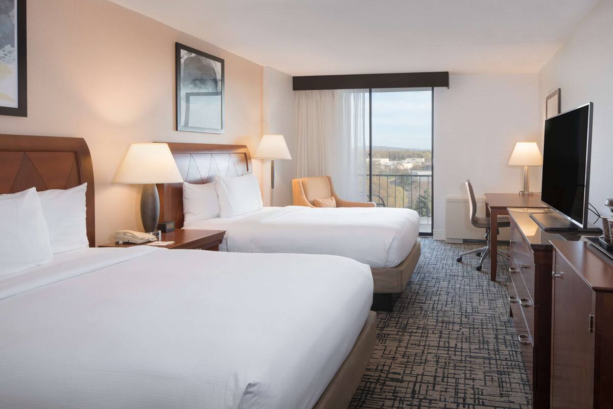 Quarto de hotel com duas camas, uma escrivaninha, uma TV e uma cadeira.