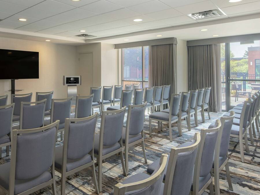 Uma sala de conferências com fileiras de cadeiras, uma TV de tela plana e grandes janelas com cortinas.