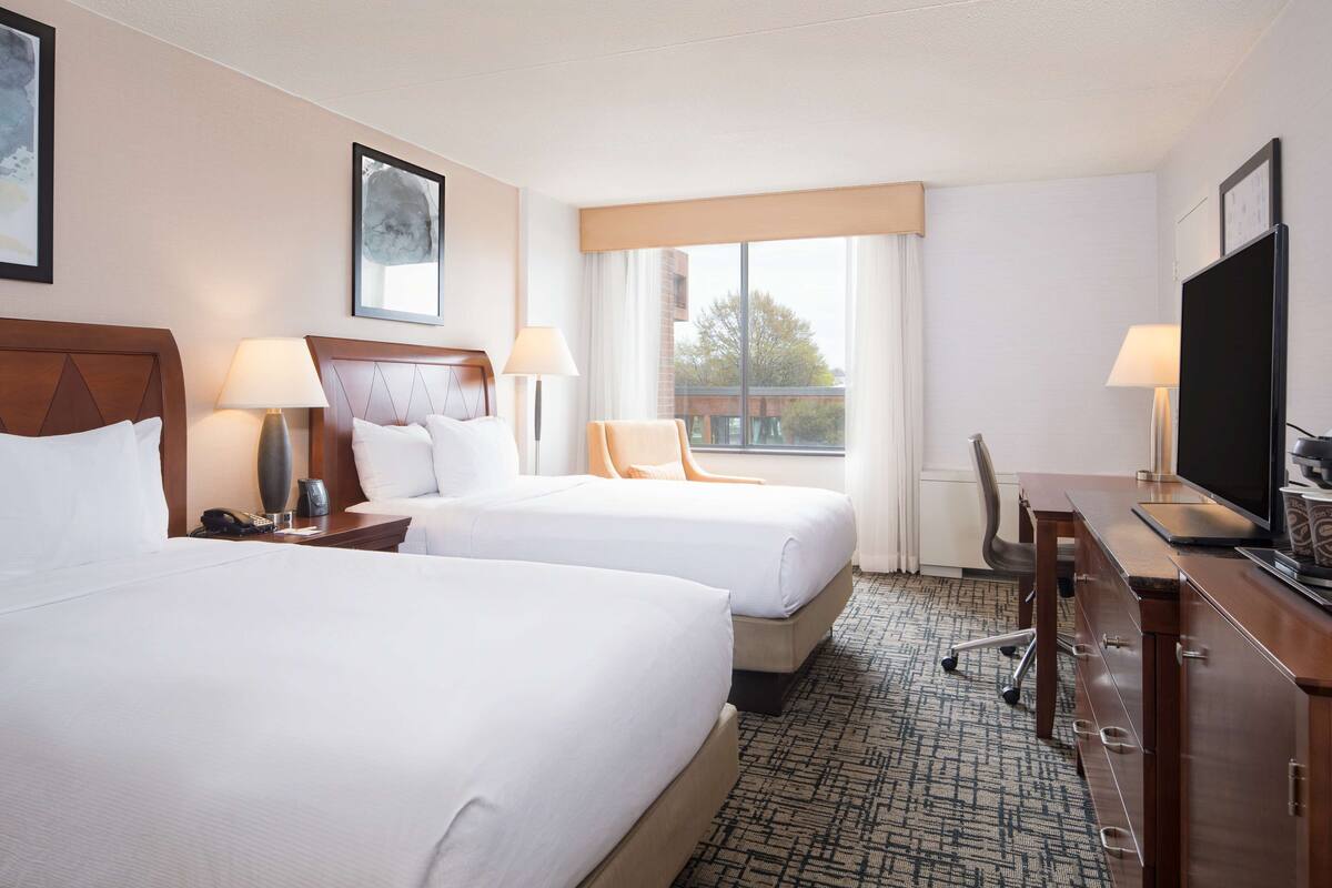 Quarto de hotel com duas camas, uma escrivaninha, uma cadeira, uma televisão e uma janela com vista para prédios.