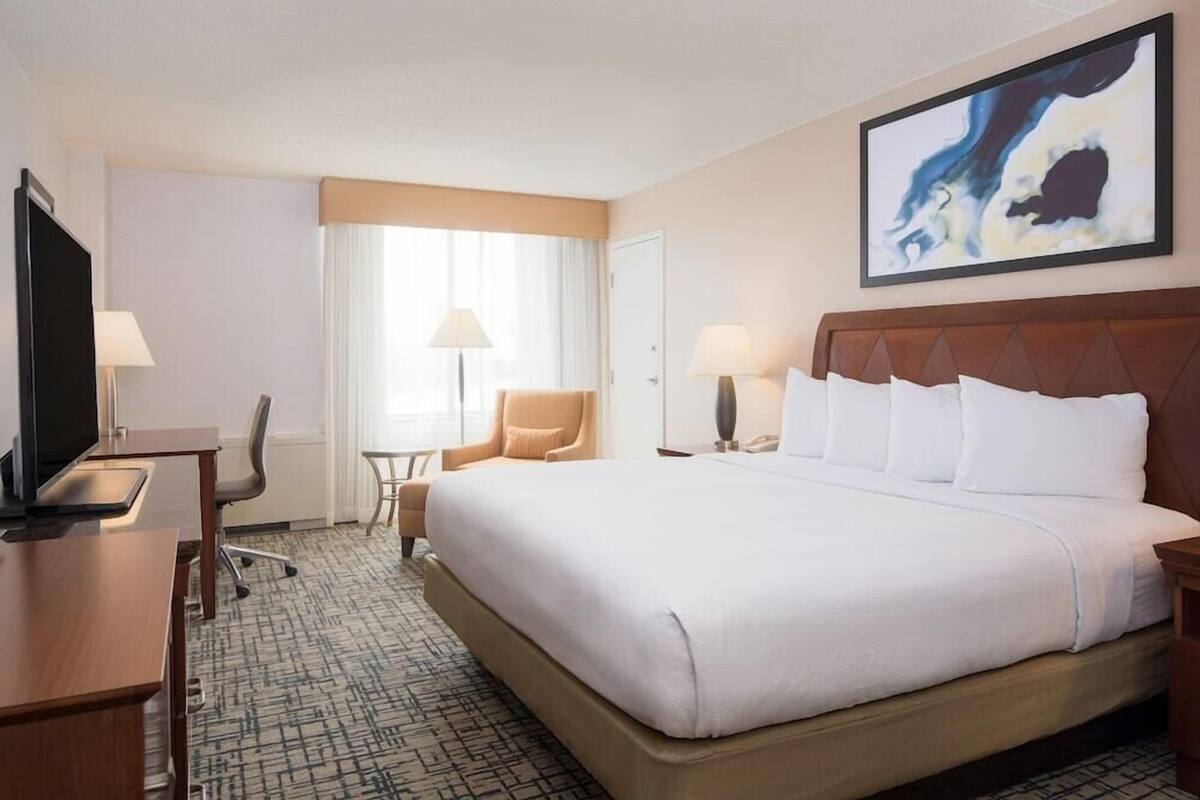 Quarto de hotel com uma cama grande, uma escrivaninha com computador, uma cadeira e uma televisão.