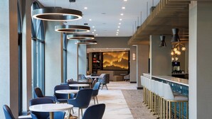 Um hall de hotel moderno com mesas redondas e cadeiras azuis, uma área de bar com banquetas e uma escadaria.