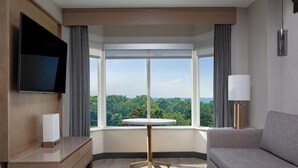 Quarto de hotel moderno com uma grande janela que oferece uma vista panorâmica, uma televisão de tela plana na parede, uma mesa redonda com base dourada e um sofá cinza.