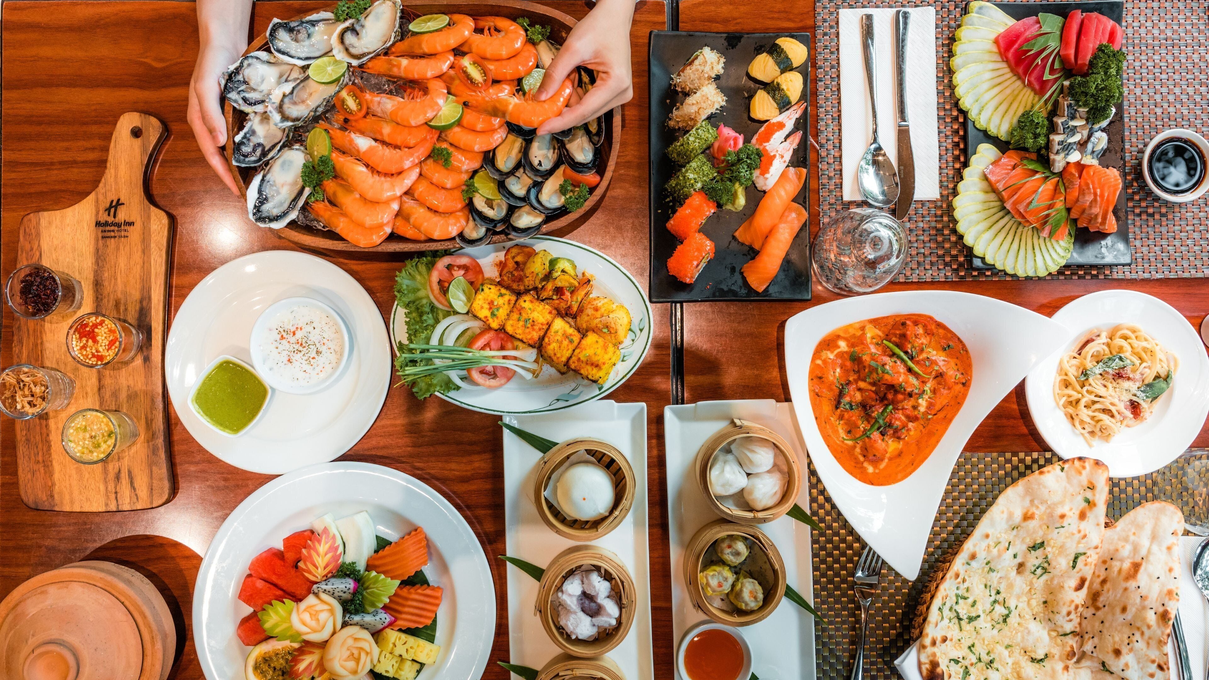 Diversos pratos, incluindo frutos do mar, sushi e massa, dispostos sobre uma mesa de madeira.