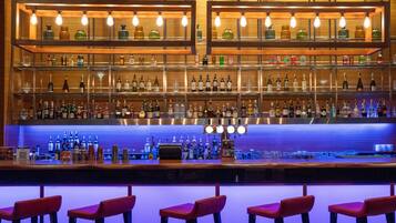 Um bar com várias prateleiras de garrafas de bebidas, um balcão com banquetas e luminárias pendentes.