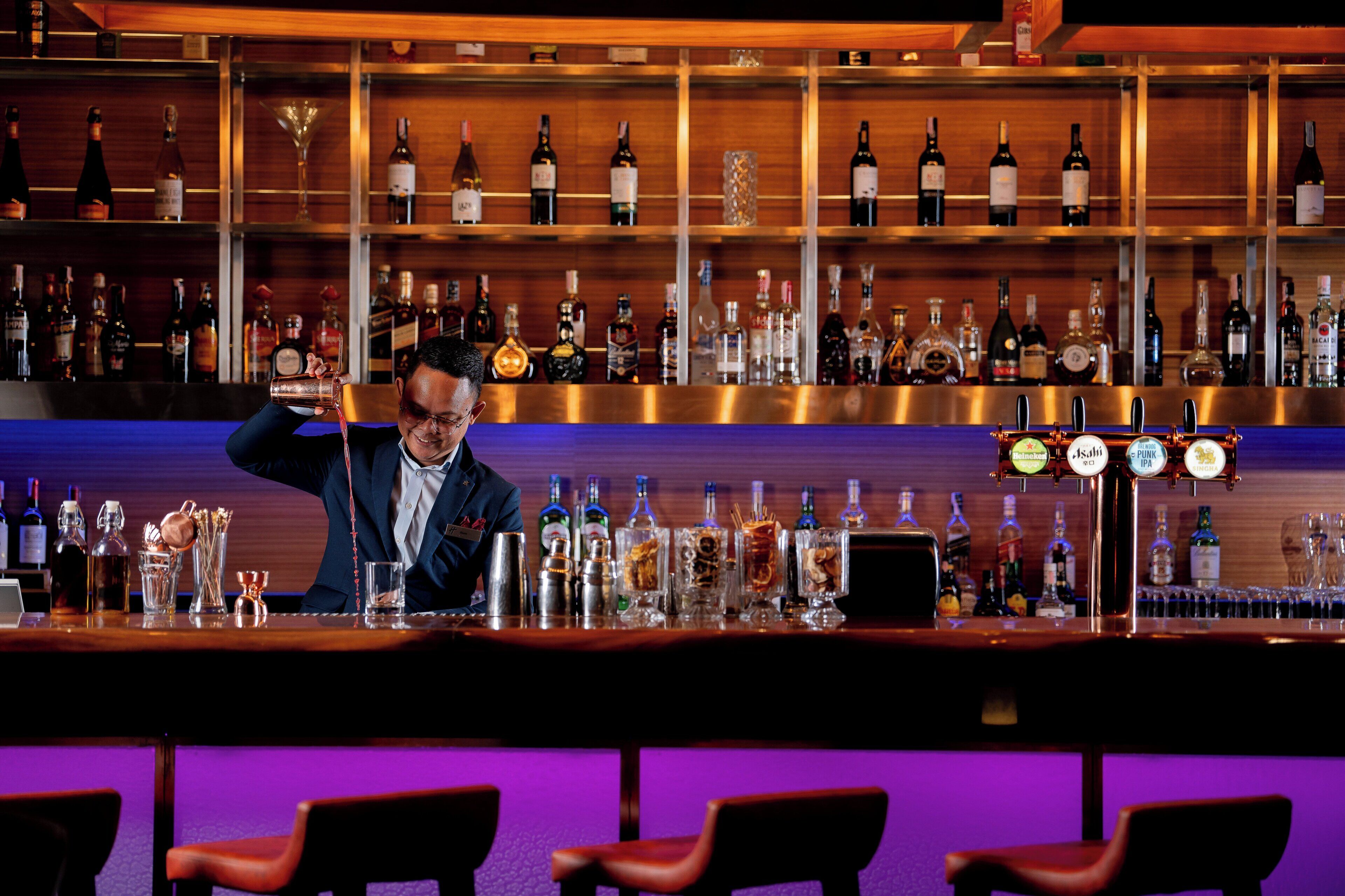 Um balcão com estantes cheias de garrafas e copos, um bartender e banquetas.