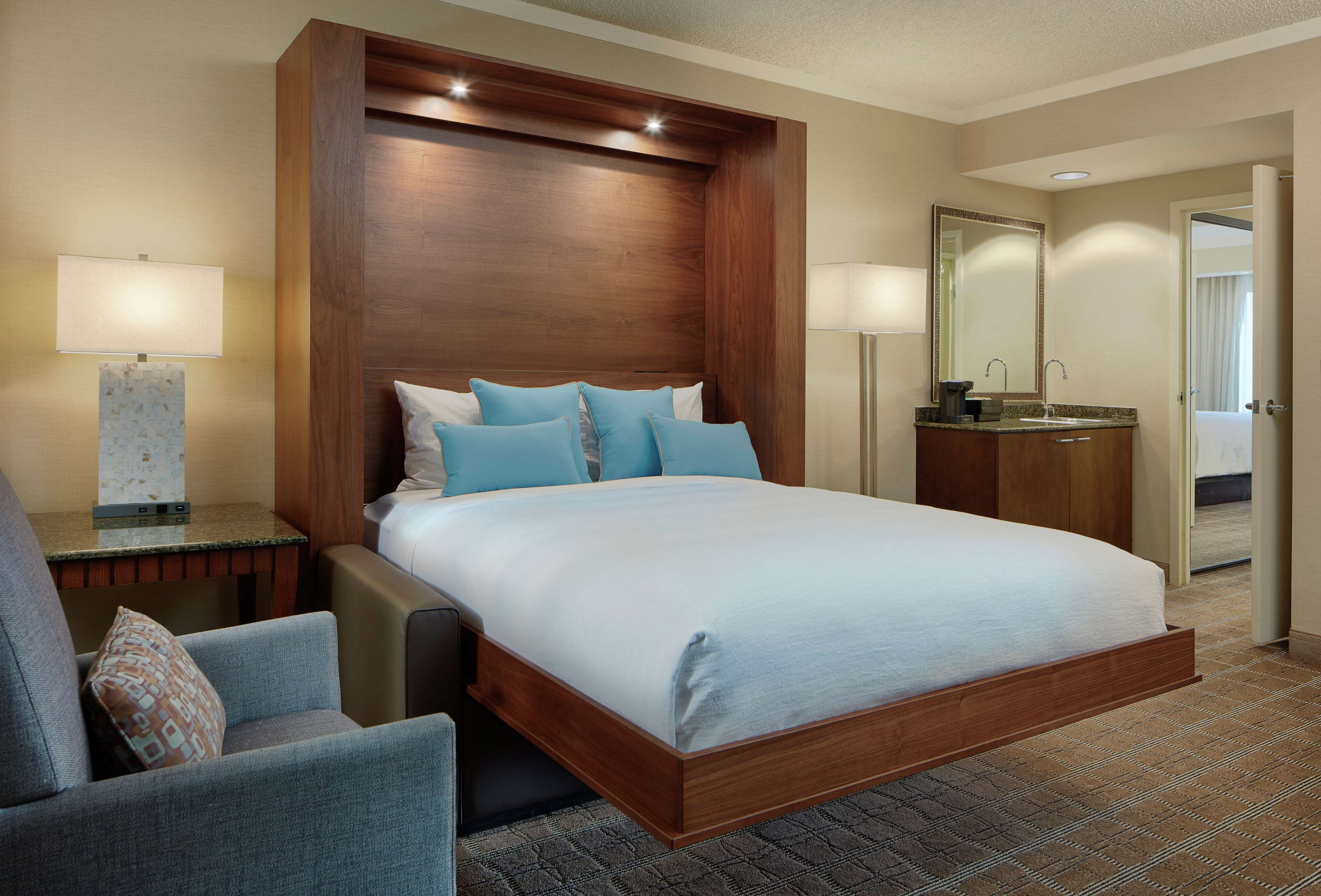 Quarto de hotel moderno com uma cama grande, cabeceira de madeira, mesa de cabeceira com abajur, uma cadeira e banheiro com espelho e pia.