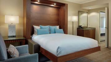 Quarto de hotel moderno com uma cama grande, cabeceira de madeira, mesa de cabeceira com abajur, uma cadeira e banheiro com espelho e pia.