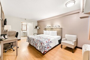 Quarto de hotel com cama, escrivaninha, cadeira, mesa de cabeceira, abajur, espelho e aquecedor fixado na parede.