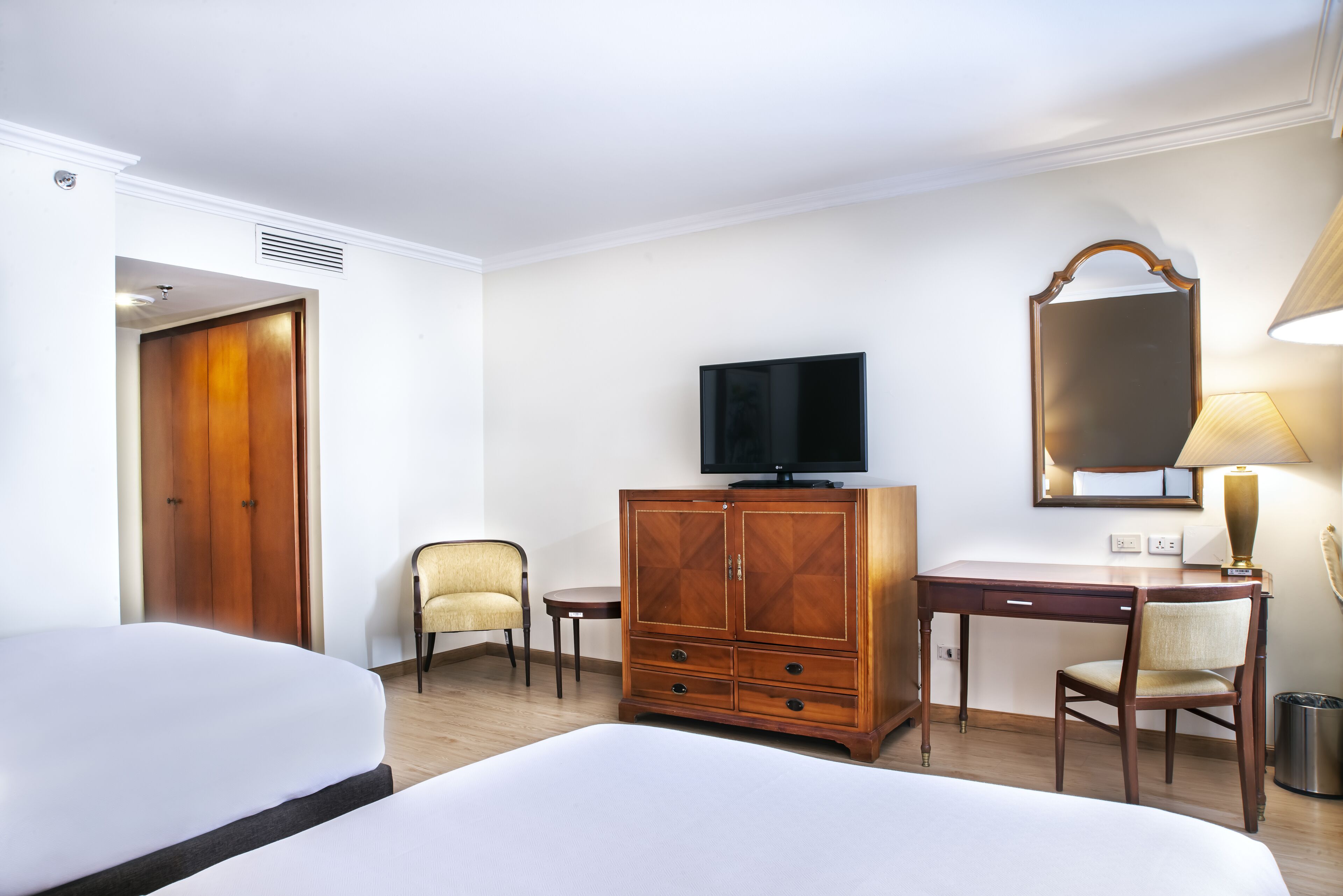 Quarto de hotel com uma cama, uma escrivaninha de madeira, uma televisão, um espelho e uma cadeira.