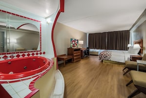 Quarto de hotel com banheira de hidromassagem vermelha, cama com colcha estampada, escrivaninha com abajur e um espelho grande.