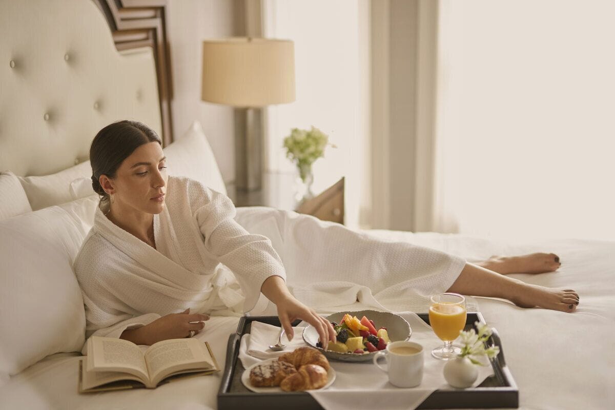 Uma pessoa deitada na cama com uma bandeja de café da manhã, incluindo croissants, frutas e suco.