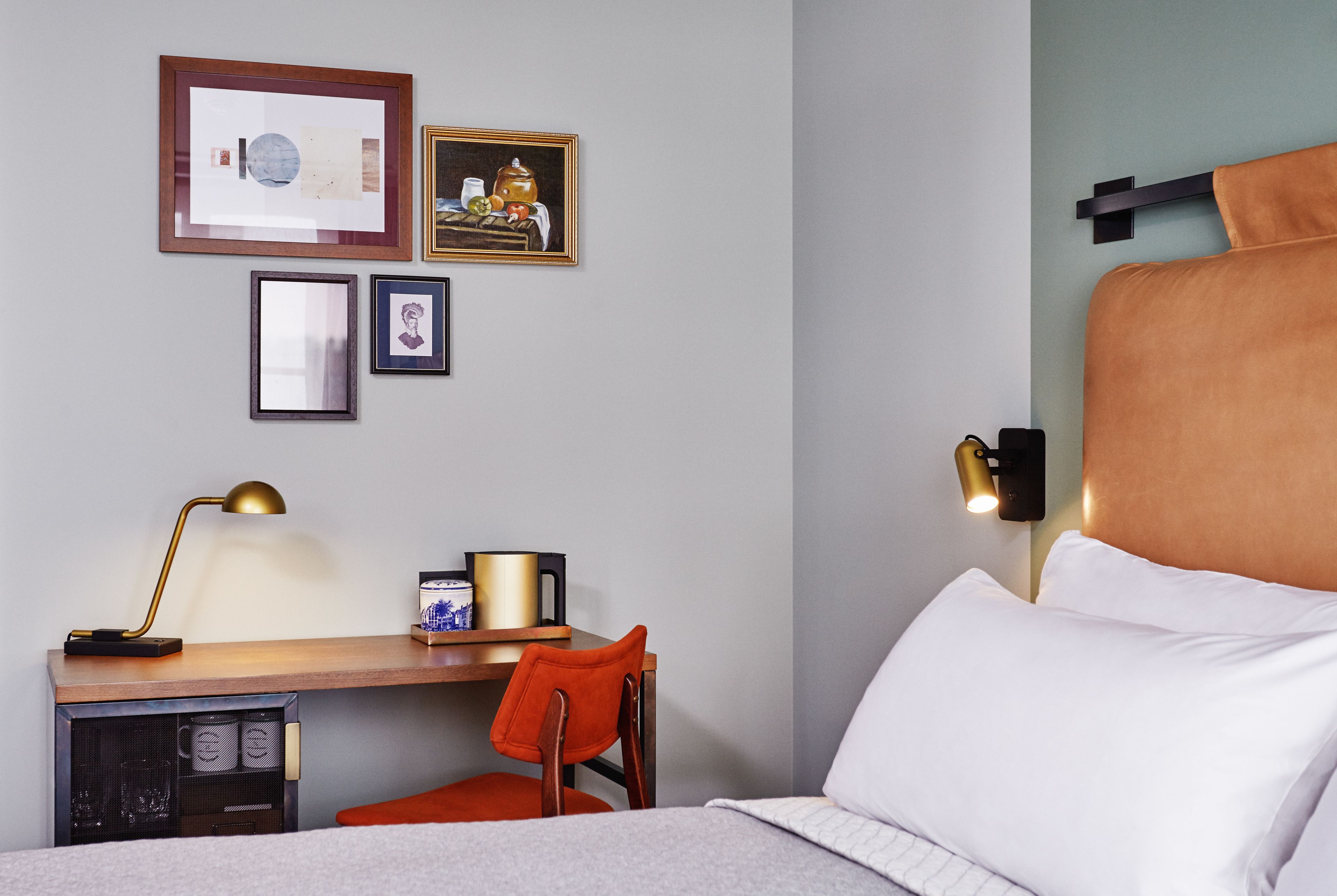 Quarto de hotel com cama, escrivaninha com luminária, cadeira, computador e obras de arte emolduradas na parede.