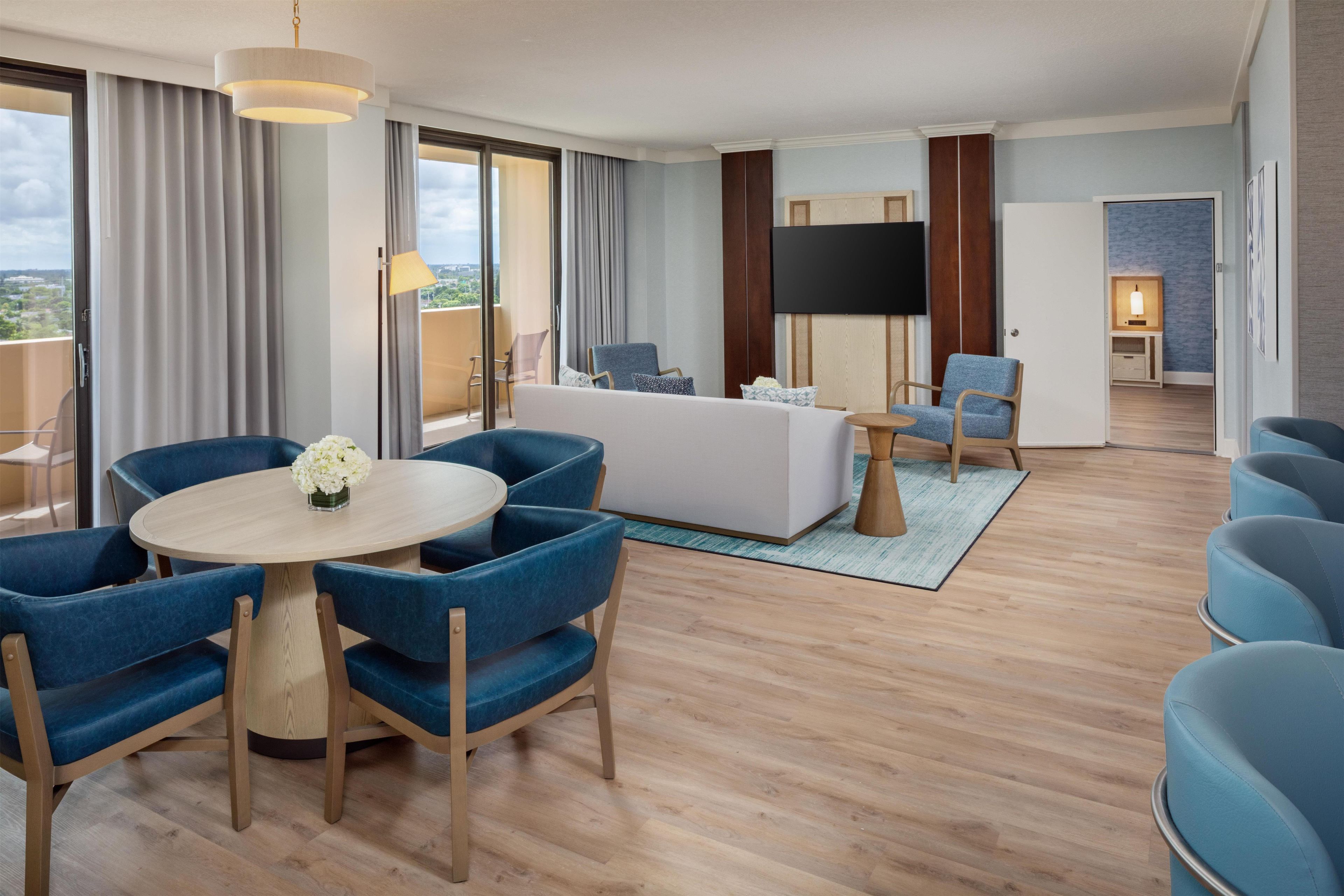 Quarto de hotel moderno com varanda, mesa de jantar redonda, cadeiras estofadas em azul, um sofá e televisão de tela plana.