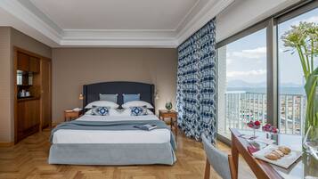 Quarto de hotel com uma cama grande, um guarda-roupa de madeira, uma pequena mesa com um copo de água e vista para a cidade através de amplas janelas.