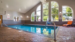 Piscina interna com vista clara para árvores através de amplas janelas em arco, uma cadeira de salva-vidas e regras da piscina afixadas na parede.