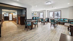 Uma sala de jantar ampla com piso de madeira e cadeiras cor turquesa, com uma cozinha ao fundo.