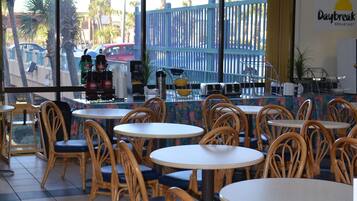 Um restaurante com cadeiras de vime e mesas redondas, vista para palmeiras e uma cerca azul do lado de fora, e uma placa com a inscrição 'Daybreak Breakfast'.