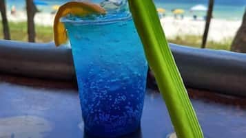 Uma bebida azul com uma fatia de laranja e um canudo verde sobre uma mesa de madeira, com vista para a praia ao fundo.