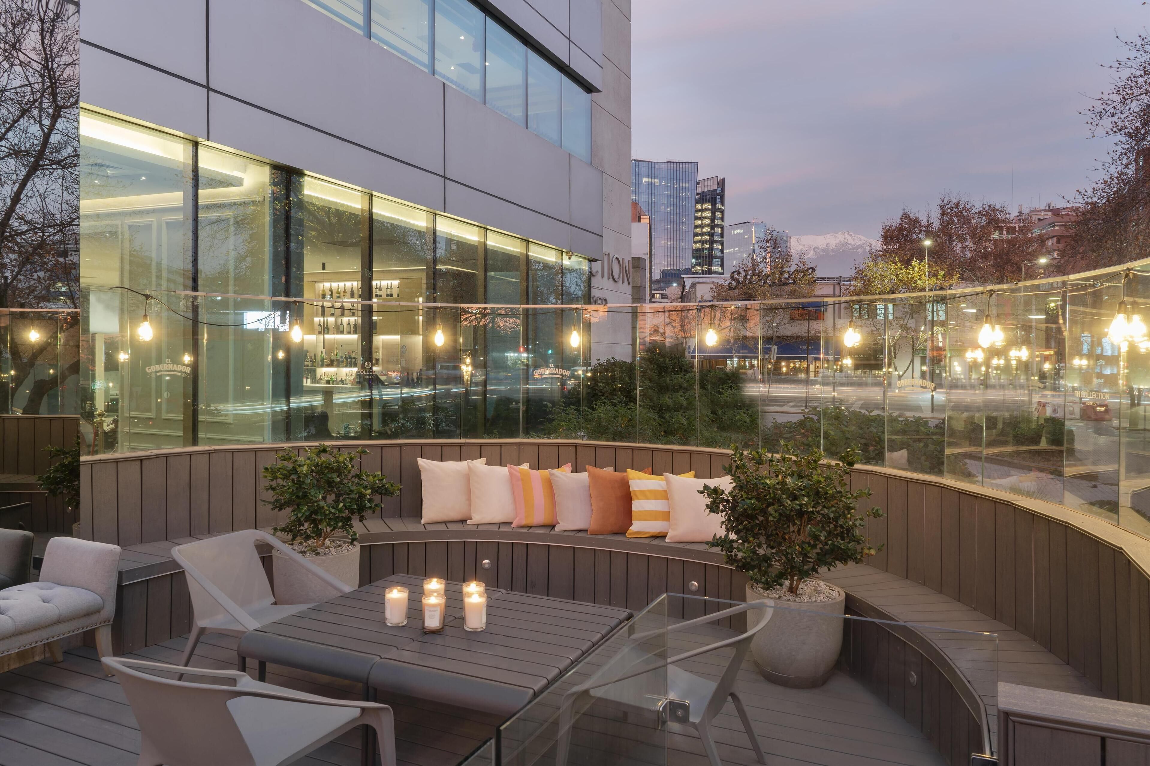 Área externa com mesas e cadeiras, vista para um edifício moderno, guarda-corpo de vidro e luzes de festa.