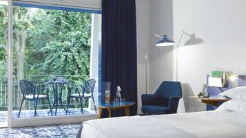 Quarto de hotel com cama, secretária com luminária, cadeira, pequena mesa com cadeiras e vista para o verde através de uma janela grande.