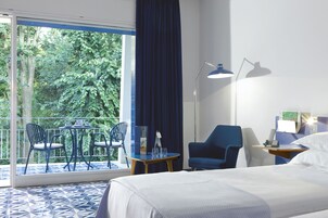 Quarto de hotel com cama, secretária com luminária, cadeira, pequena mesa com cadeiras e vista para o verde através de uma janela grande.