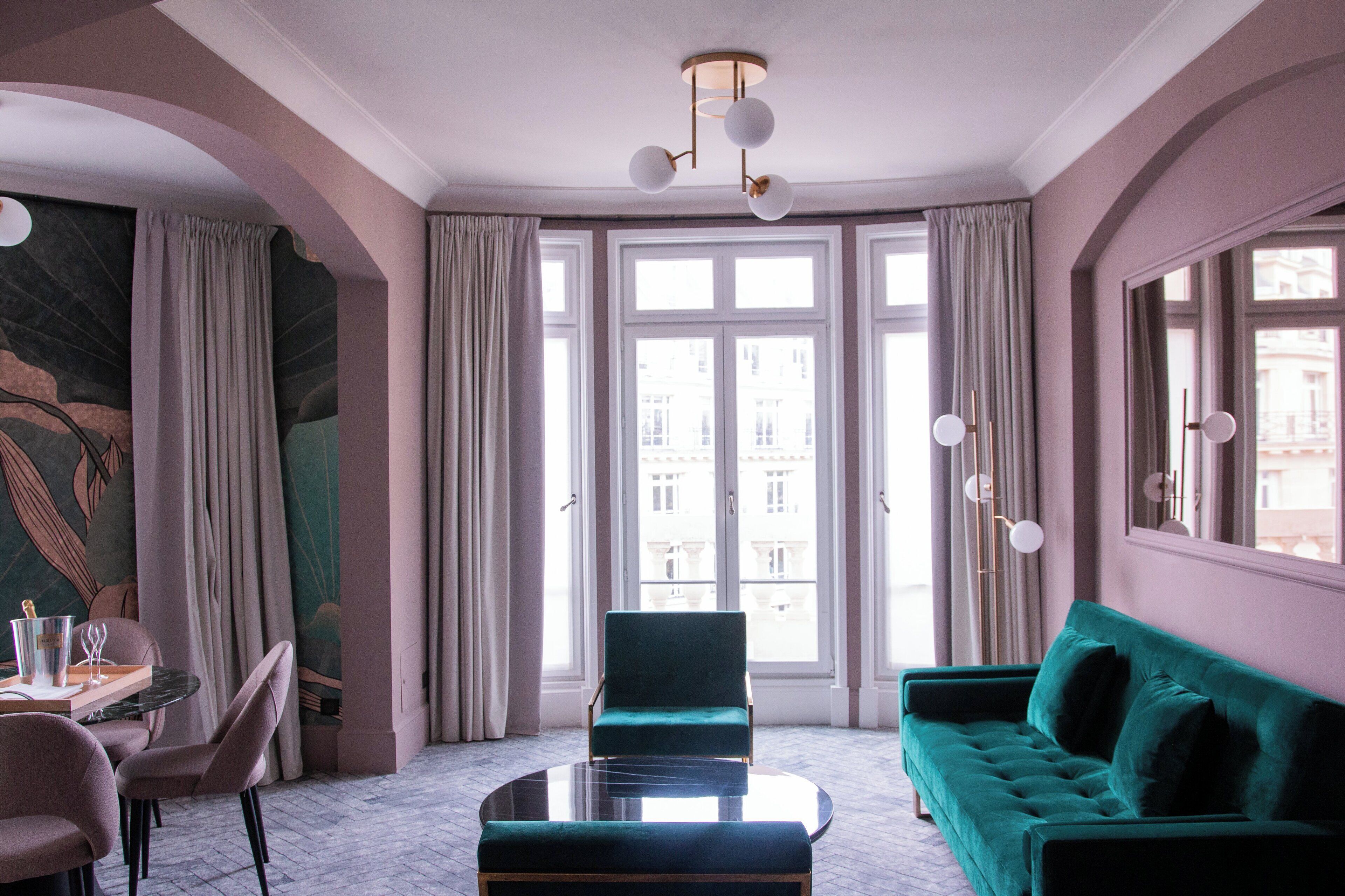 Quarto de hotel moderno com sofá verde, mesa redonda com cadeiras e janelas amplas com cortinas.