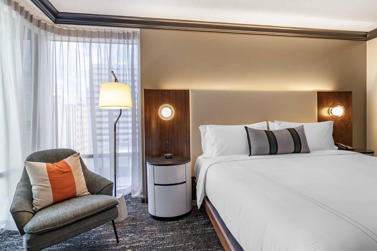 Quarto de hotel com uma cama grande, uma cadeira com almofada, mesa de cabeceira com abajur e vista para a cidade pela janela.