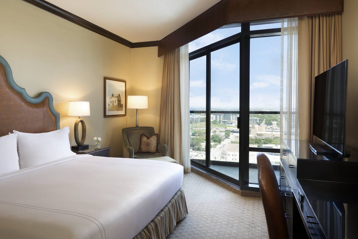 Quarto de hotel com uma cama grande, uma escrivaninha, uma cadeira, uma TV e uma janela ampla com vista para a cidade.