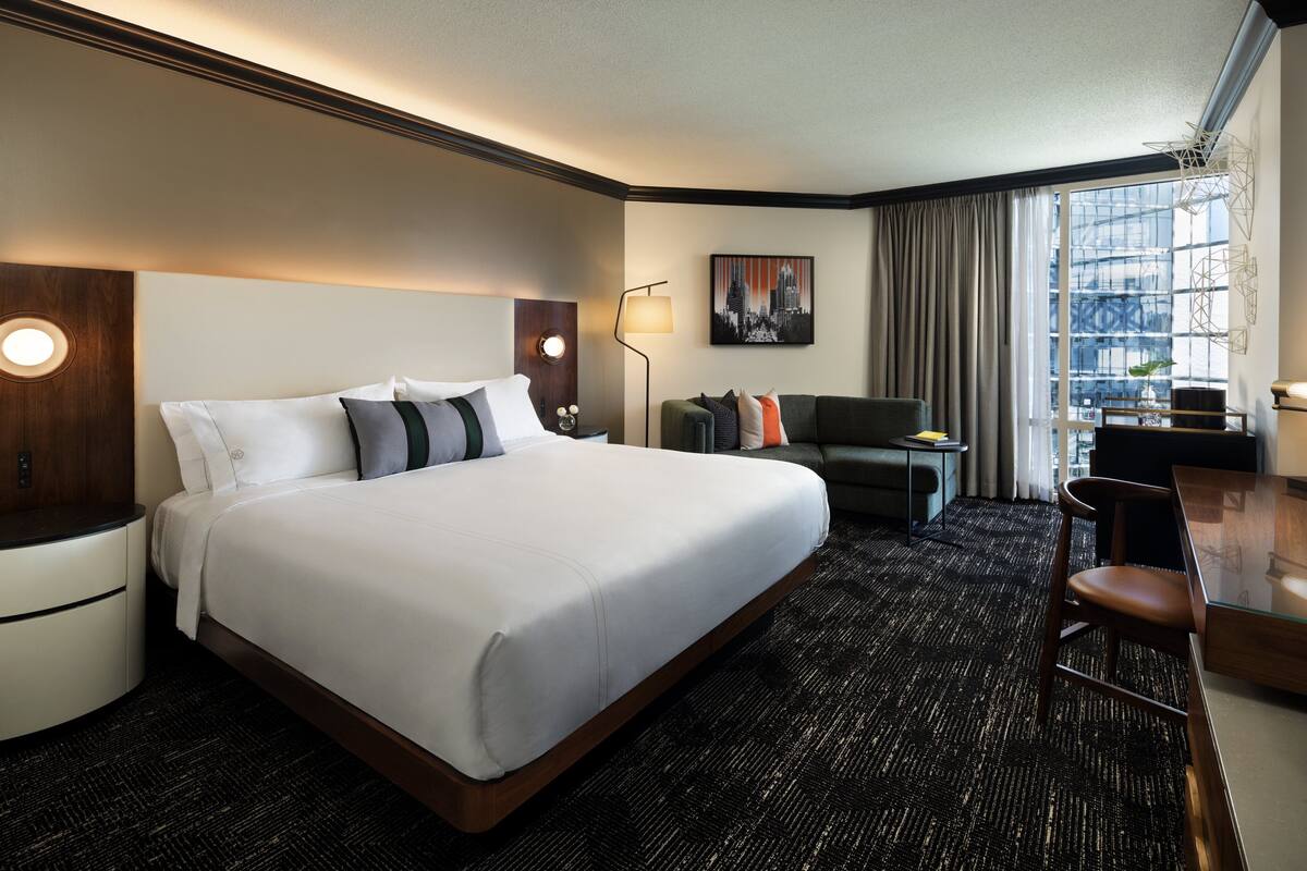 Quarto de hotel com uma cama grande, uma escrivaninha, uma cadeira, um abajur e um quadro na parede.
