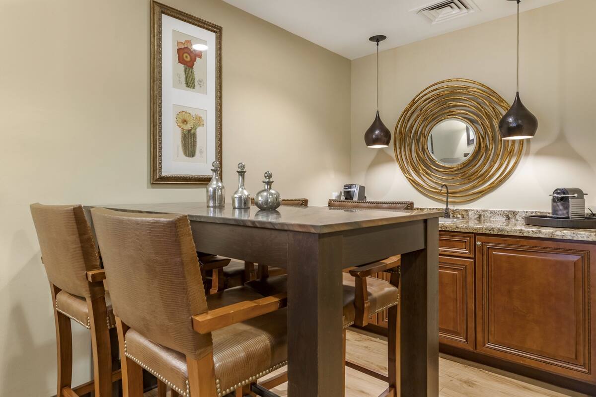 Uma área de refeições com mesa e cadeiras de madeira, uma ilustração botânica emoldurada, luminárias pendentes e uma bancada de cozinha com armários.