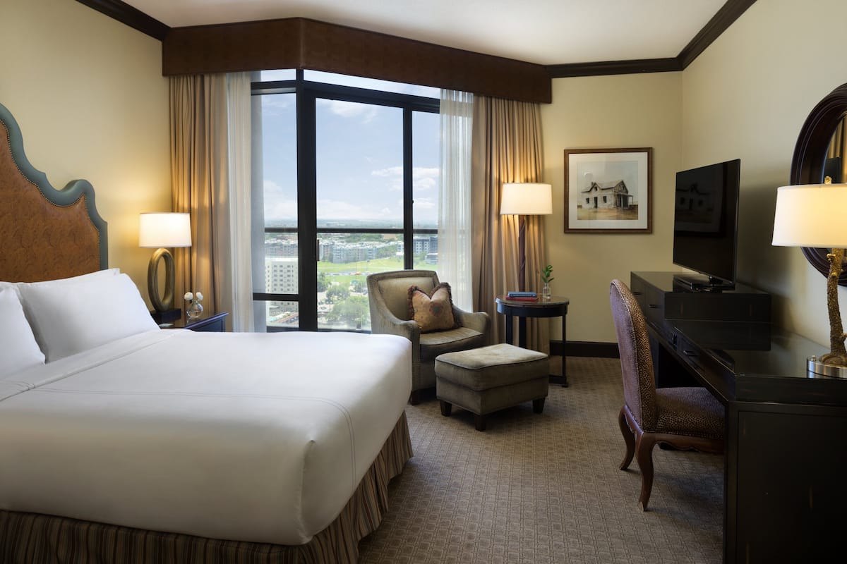 Quarto de hotel com uma cama grande, uma mesa de escritório, uma cadeira, uma mesinha e vista para a cidade através de amplas janelas.