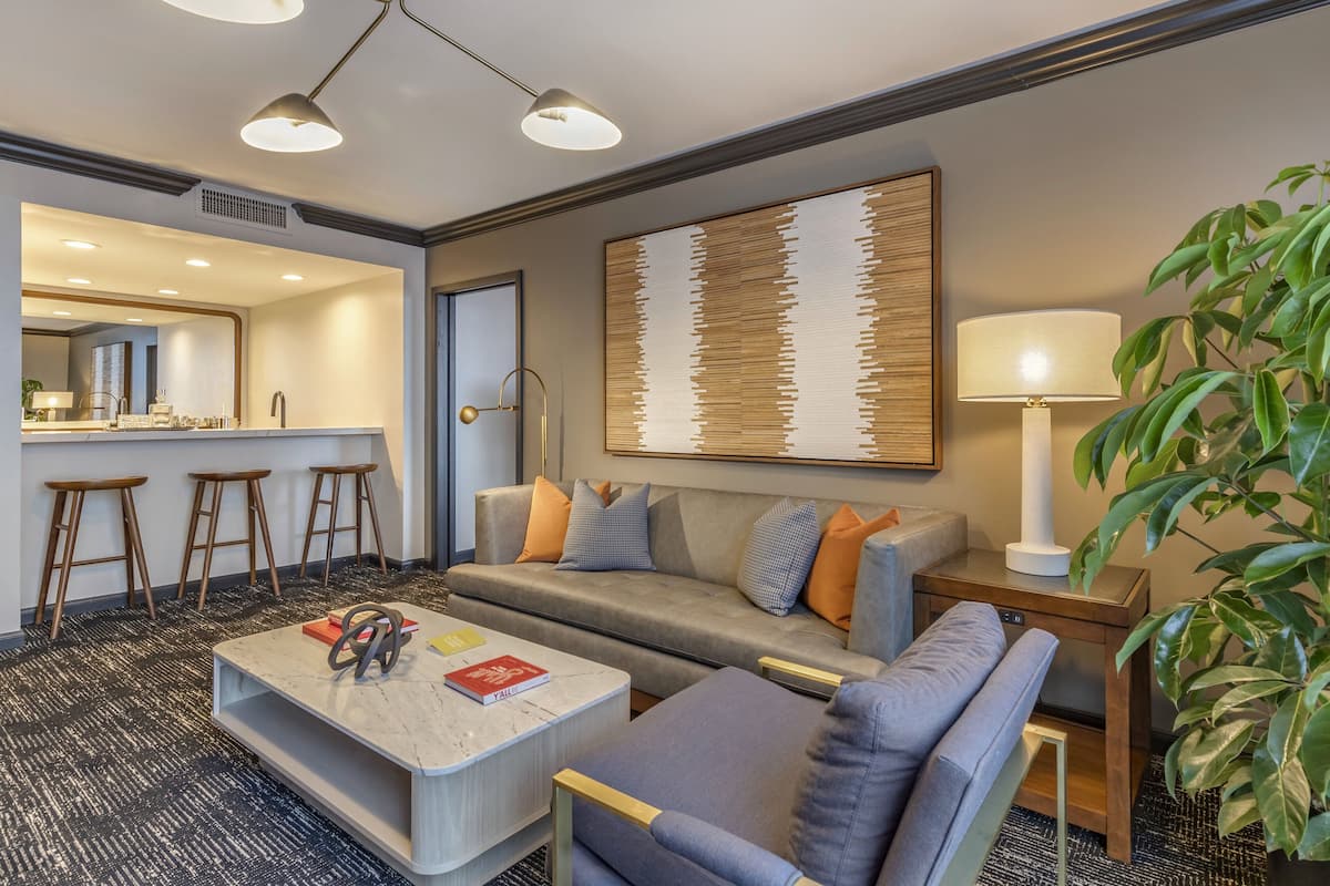 Quarto de hotel moderno com sofá, poltronas, mesa de centro, abajur e planta.