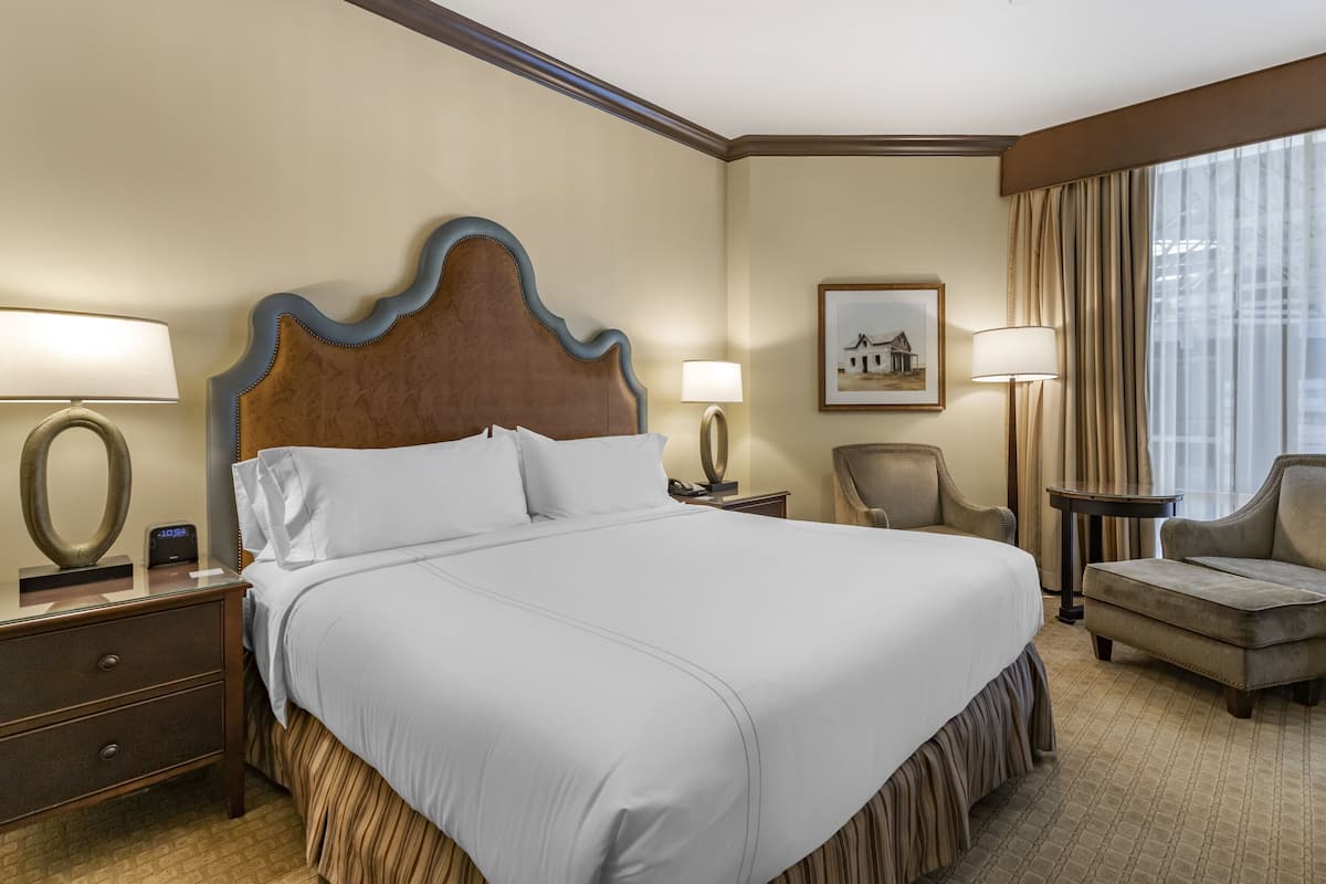 Quarto de hotel com uma cama grande, uma cadeira, uma mesinha de cabeceira, um abajur e um quadro na parede.