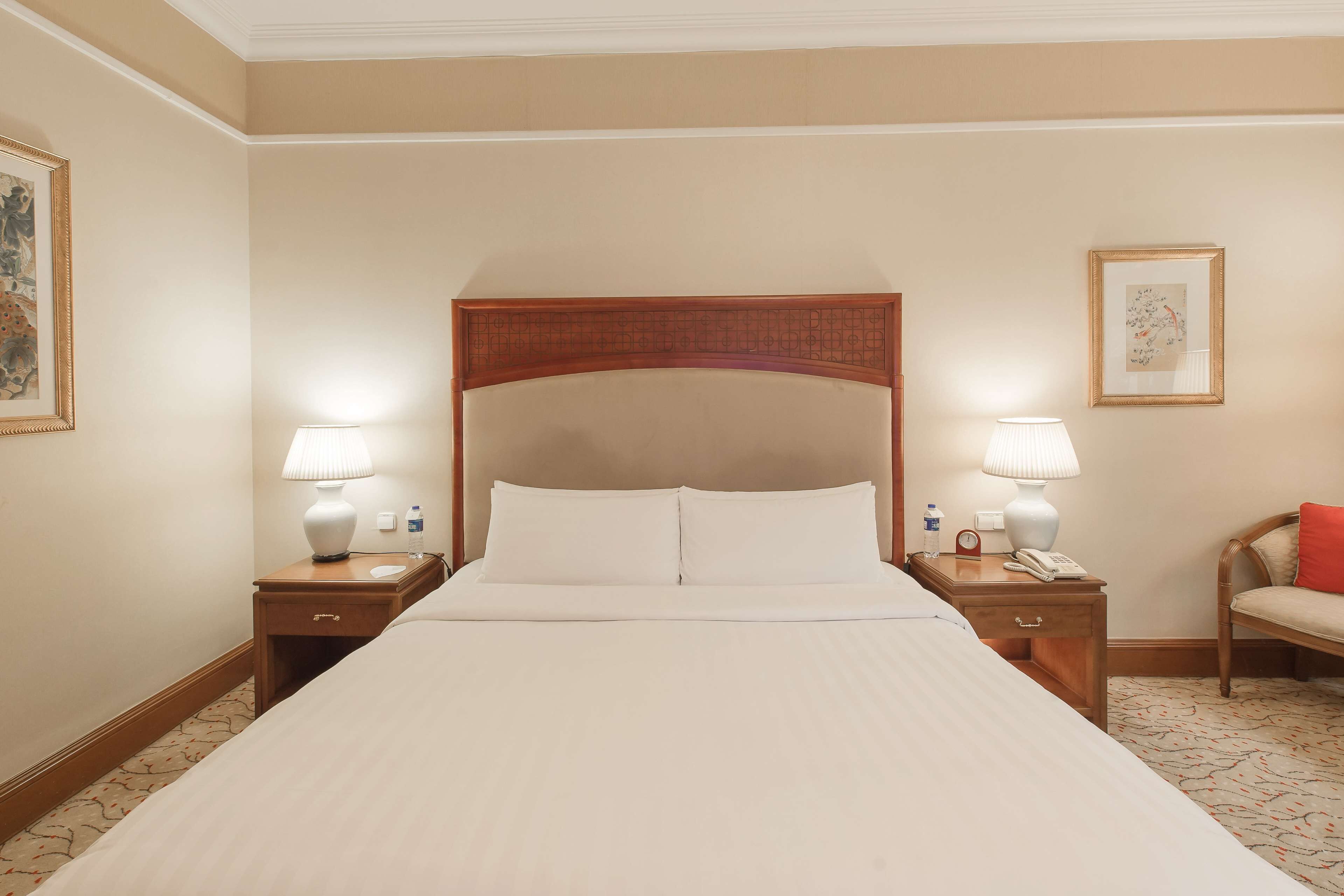 Quarto de hotel com uma cama grande, duas mesas de cabeceira com abajures, uma cadeira e um quadro na parede.