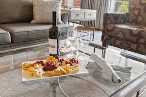 Uma mesa com tampo de vidro, uma garrafa de vinho, duas taças, um tabuleiro de queijos, bolachas e uvas.