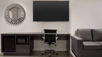 Uma sala de estar moderna com um console de madeira escura, uma cadeira de escritório preta, um sofá cinza, uma grande televisão de tela plana e um espelho circular único.