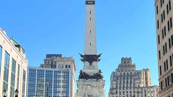 Um monumento alto com uma estátua no topo, rodeado por edifícios modernos e carros estacionados.