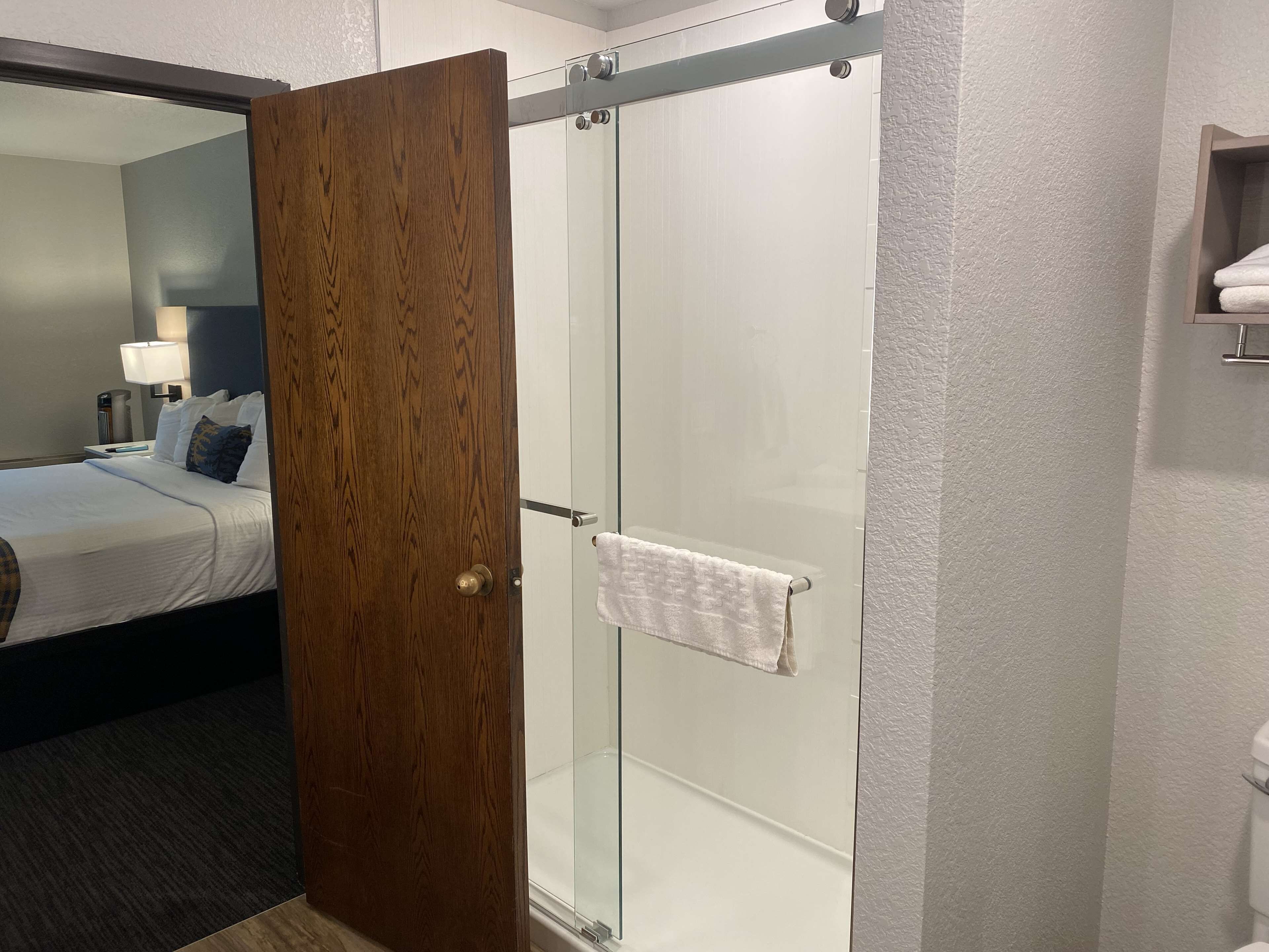 Quarto de hotel com porta de madeira, box de vidro, cama com roupa de cama branca e mesa de cabeceira com luminária.