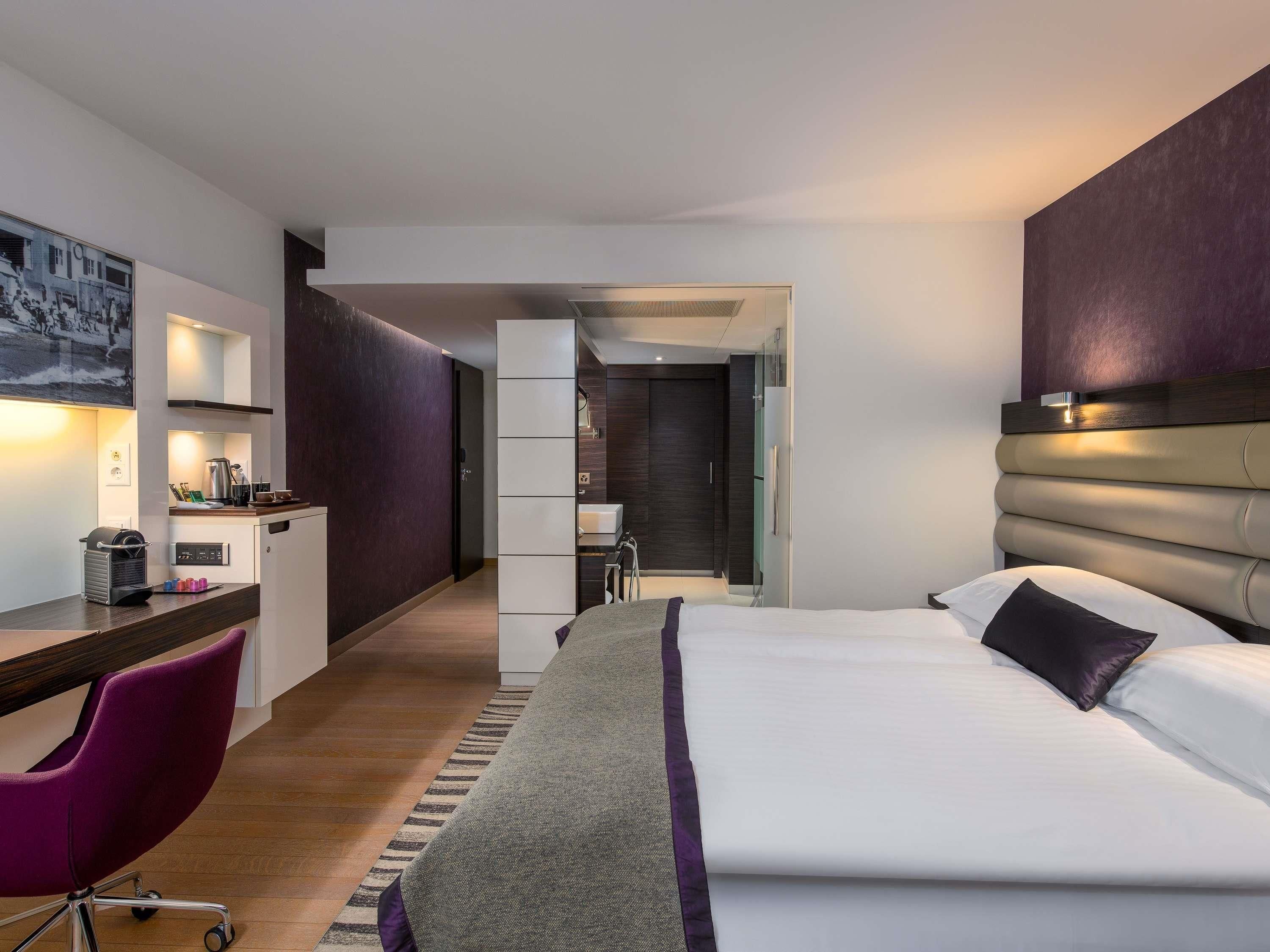 Quarto de hotel moderno com uma cama grande, uma escrivaninha com cadeira, uma cozinha compacta e um banheiro visível através de uma porta aberta.